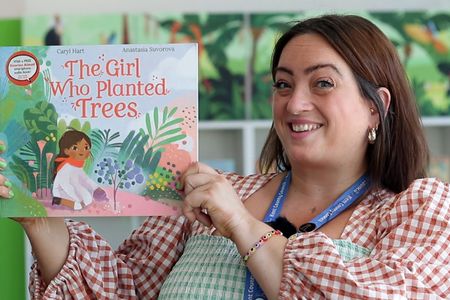 A staff member from Newington Library holding up a book called "The Girl Who Planted Trees" and smiling 